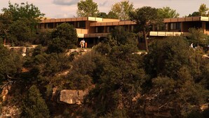 Um edifício moderno com telhado plano e fachada de tijolo e vidro, cercado por vegetação densa e terreno rochoso.