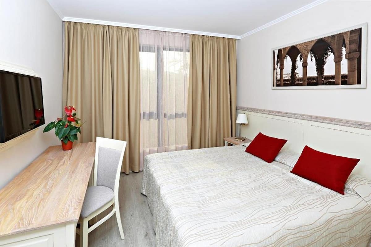 Quarto de hotel com cama, escrivaninha, cadeira, televisão, luminária e um quadro na parede.