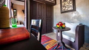 Quarto com sofá, mesinha redonda com arranjo de flores, cadeira, cesto de vime e um copo de vidro verde.