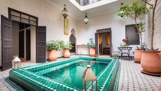 Um pátio com piscina em mosaico, plantas em vasos e área de estar com mesa e cadeiras.