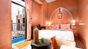Quarto de hotel com cama, luminárias de cabeceira, uma mesinha com um vaso e vista para a varanda externa.