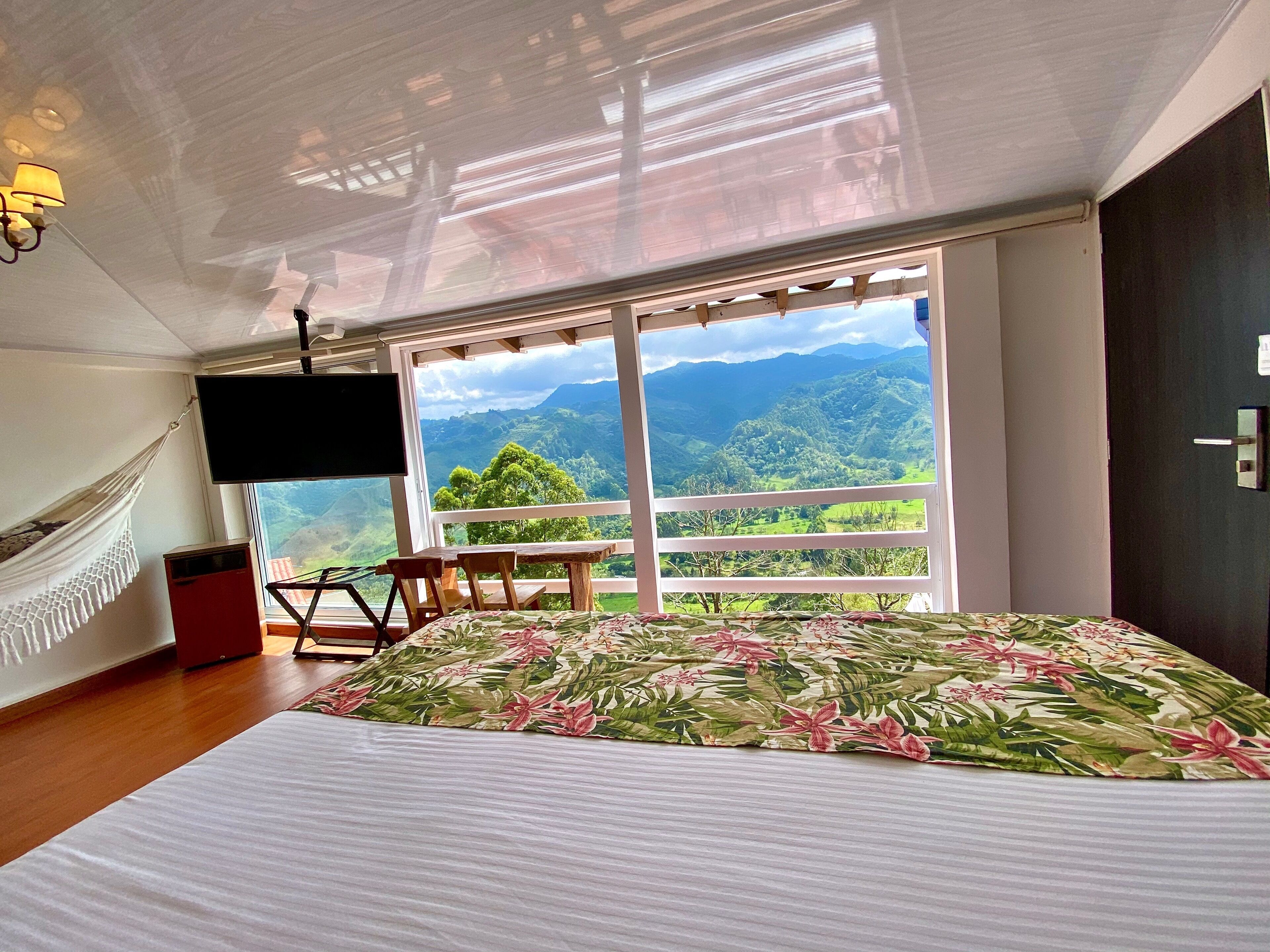 Quarto com cama grande, televisão, rede e vista para montanhas através de uma janela ampla.