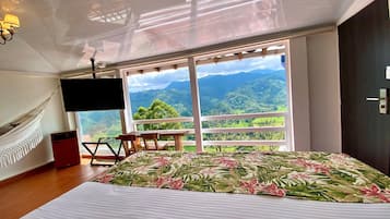 Quarto com cama grande, televisão, rede e vista para montanhas através de uma janela ampla.
