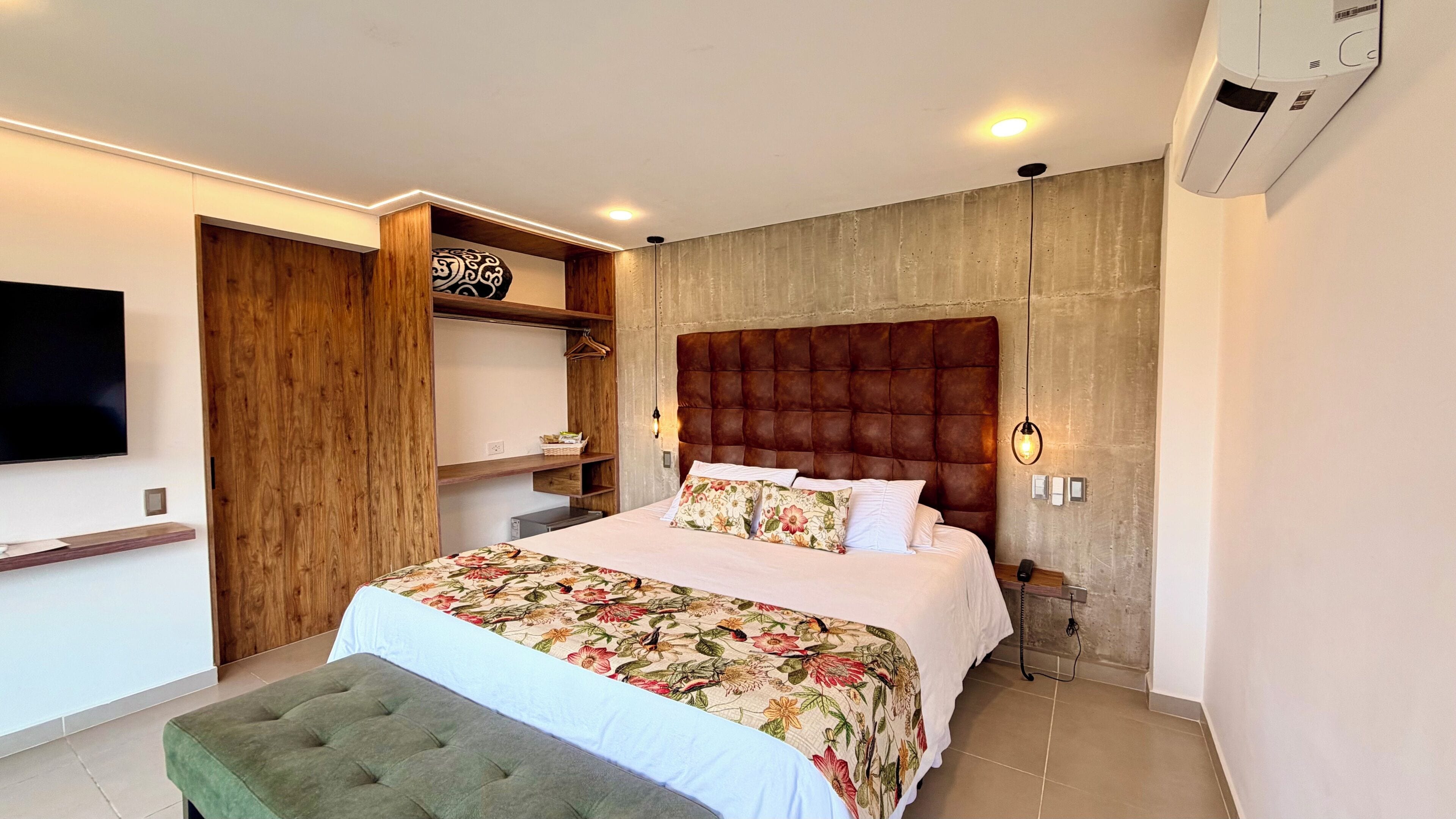 Quarto com cama, televisão, guarda-roupa, um banco e ar condicionado fixado na parede.