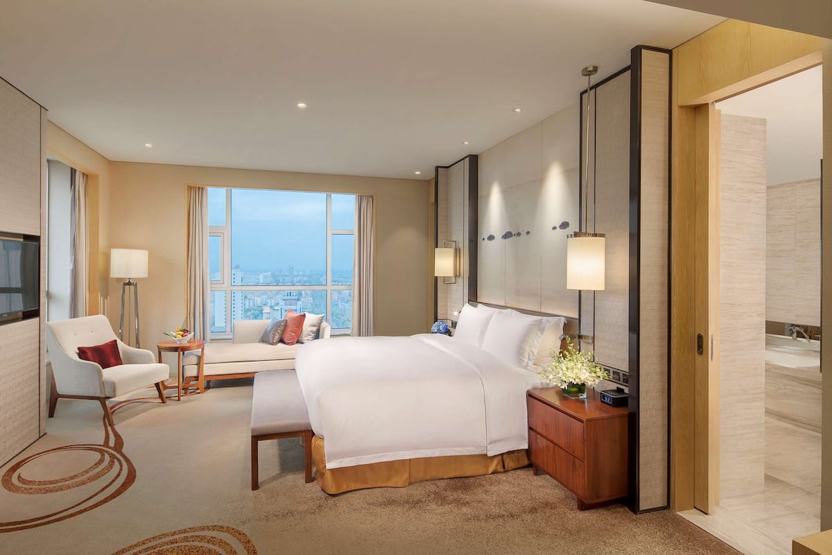 Quarto de hotel com cama grande, área de estar com cadeira e mesa, TV e vista para a cidade.
