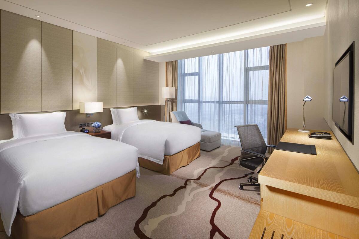 Quarto de hotel com duas camas, uma escrivaninha com cadeira, televisão e uma janela grande com cortinas.