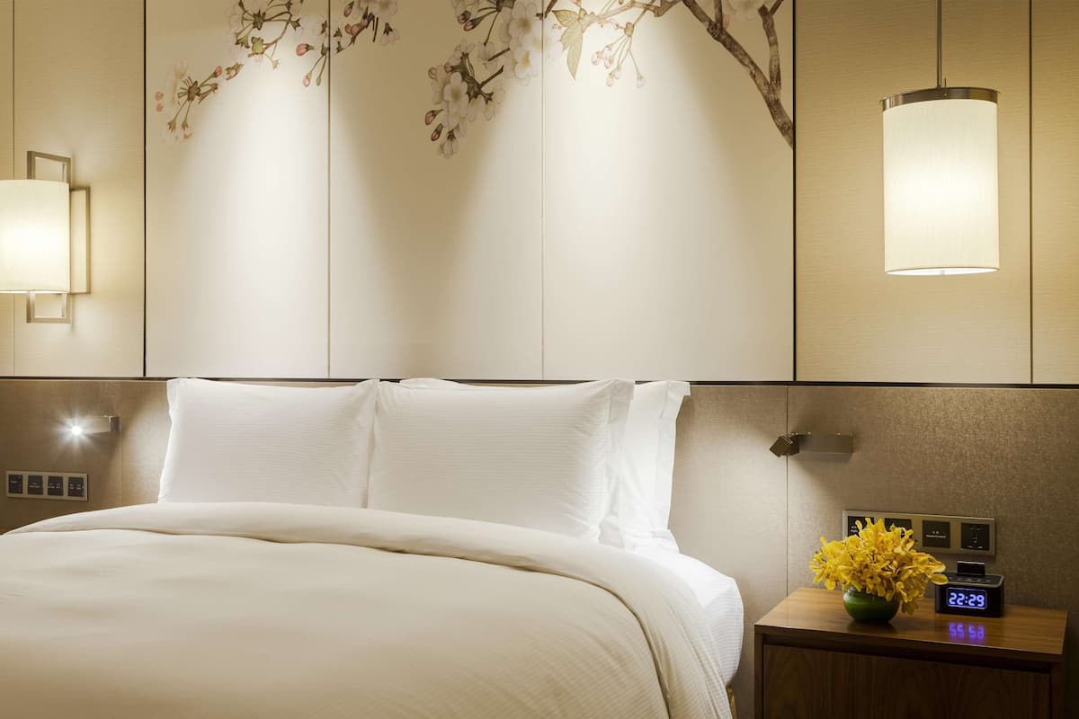 Quarto de hotel com uma cama grande, duas luminárias fixadas na parede, um relógio e um vaso com flores sobre uma mesa de cabeceira.