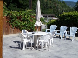 Um pátio com uma mesa redonda e seis cadeiras de plástico brancas, um guarda-sol e uma cerca de madeira com folhagem verde.