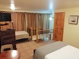 Quarto de hotel com duas camas, uma escrivaninha, uma cadeira, televisão e varanda com guarda-corpo.