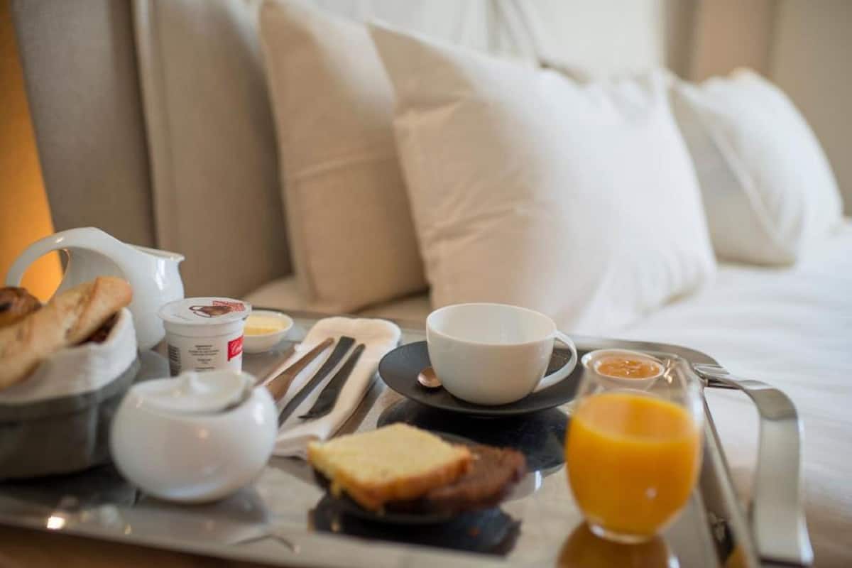 Bandeja de café da manhã com torradas, manteiga, geleia, uma xícara e suco de laranja sobre uma cama com travesseiros brancos.