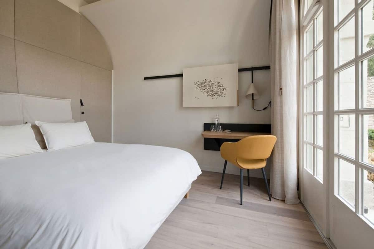 Quarto de hotel moderno com uma cama grande, uma escrivaninha, uma cadeira, uma luminária na parede e uma janela com cortinas.