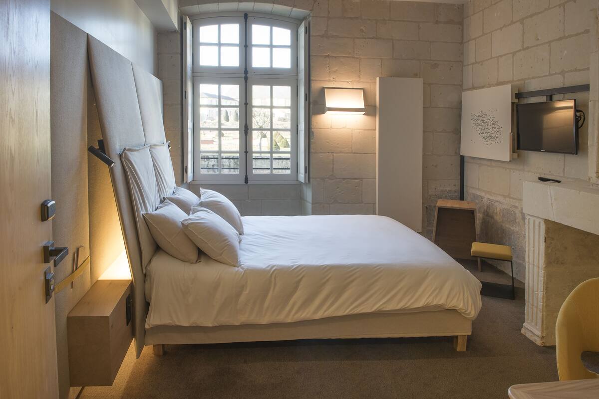 Quarto com cama grande, janela, televisão fixada na parede e mesa de cabeceira.