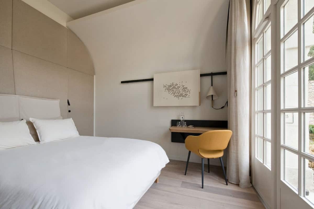 Quarto de hotel moderno com uma cama grande, uma escrivaninha e uma cadeira. Há uma janela com cortinas e uma obra de arte pendurada na parede.