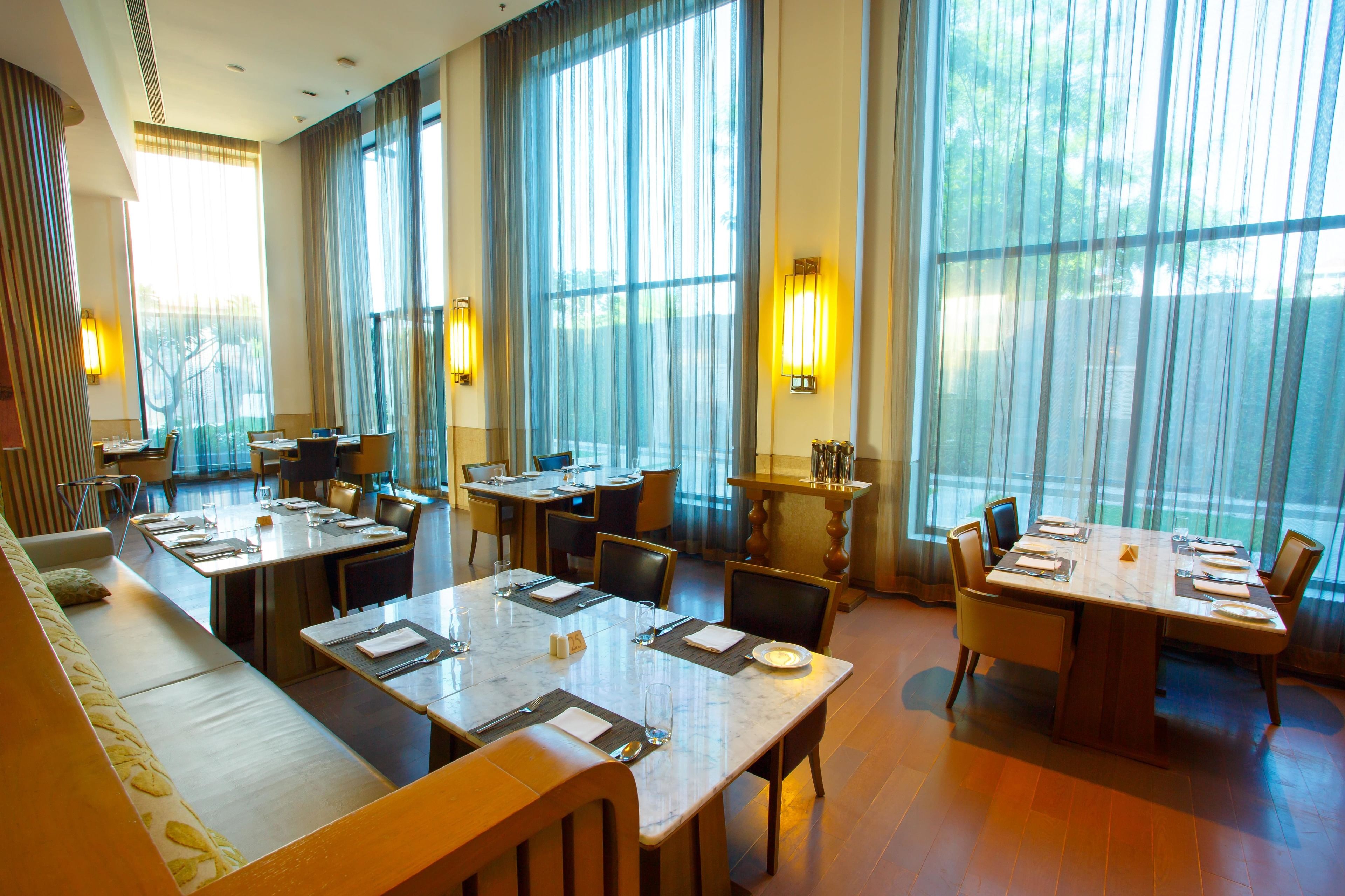 Uma sala de jantar de hotel com várias mesas e cadeiras, janelas amplas com cortinas e iluminação embutida nas paredes.
