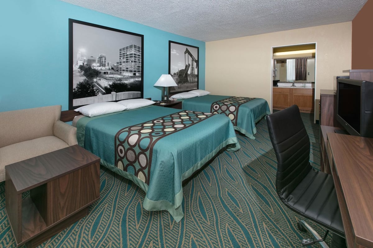 Quarto de hotel com duas camas, uma mesa, uma cadeira, uma televisão e um banheiro visível por uma porta aberta.