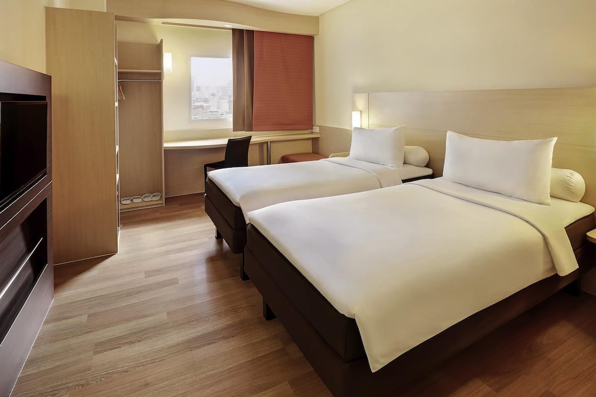 Quarto de hotel com duas camas, uma escrivaninha, uma cadeira, uma televisão e janela com vista para a cidade.