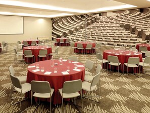 Sala de conferências com mesas e cadeiras redondas, piso coberto por carpete e um amplo auditório ao fundo.