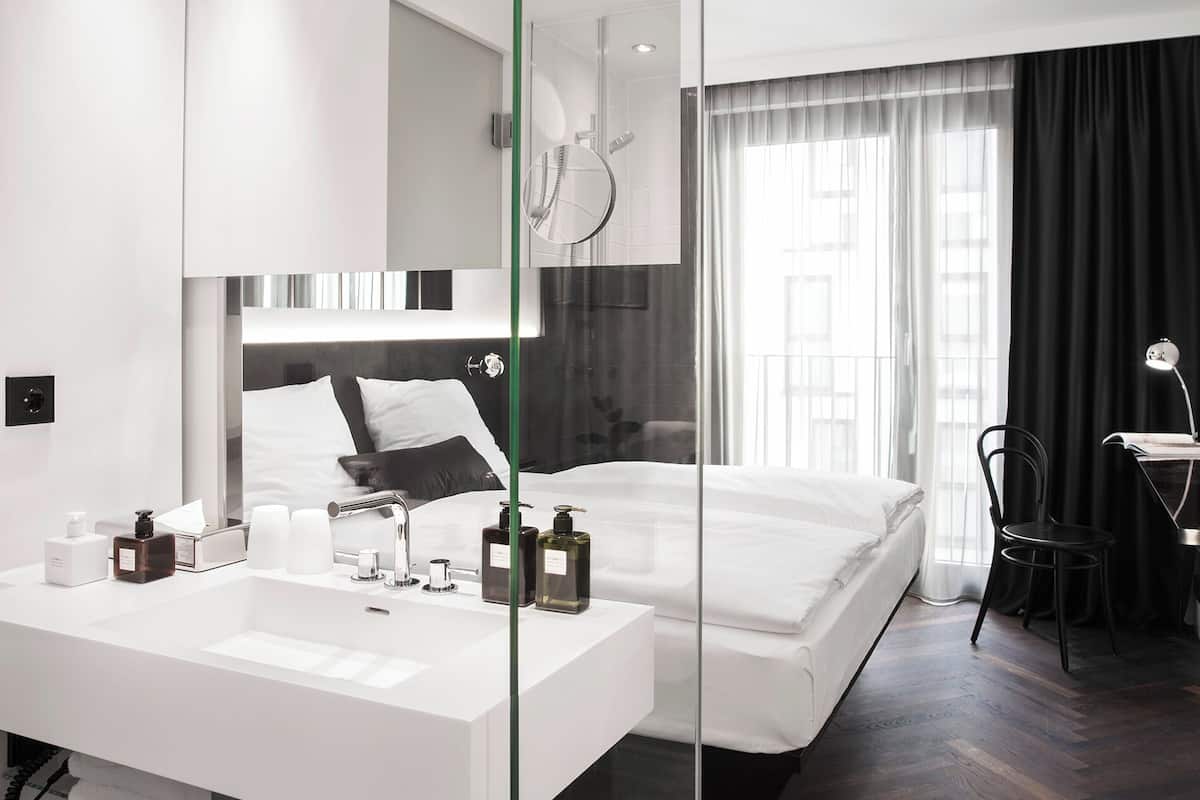 Quarto de hotel moderno com uma cama grande, chuveiro em cabine de vidro, toucador branco com artigos de higiene e vista da cidade por grandes janelas.