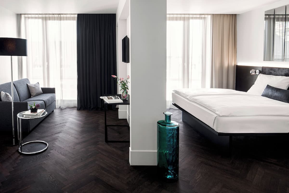 Quarto de hotel moderno com uma cama, um sofá, uma mesa de trabalho e uma janela grande.