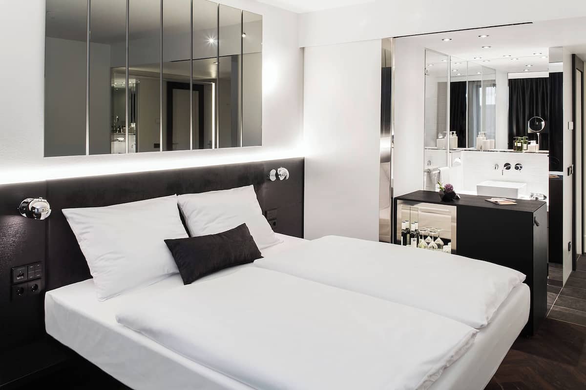 Quarto de hotel moderno com uma cama grande, mesa de cabeceira, espelho e banheiro visível através de uma porta aberta.