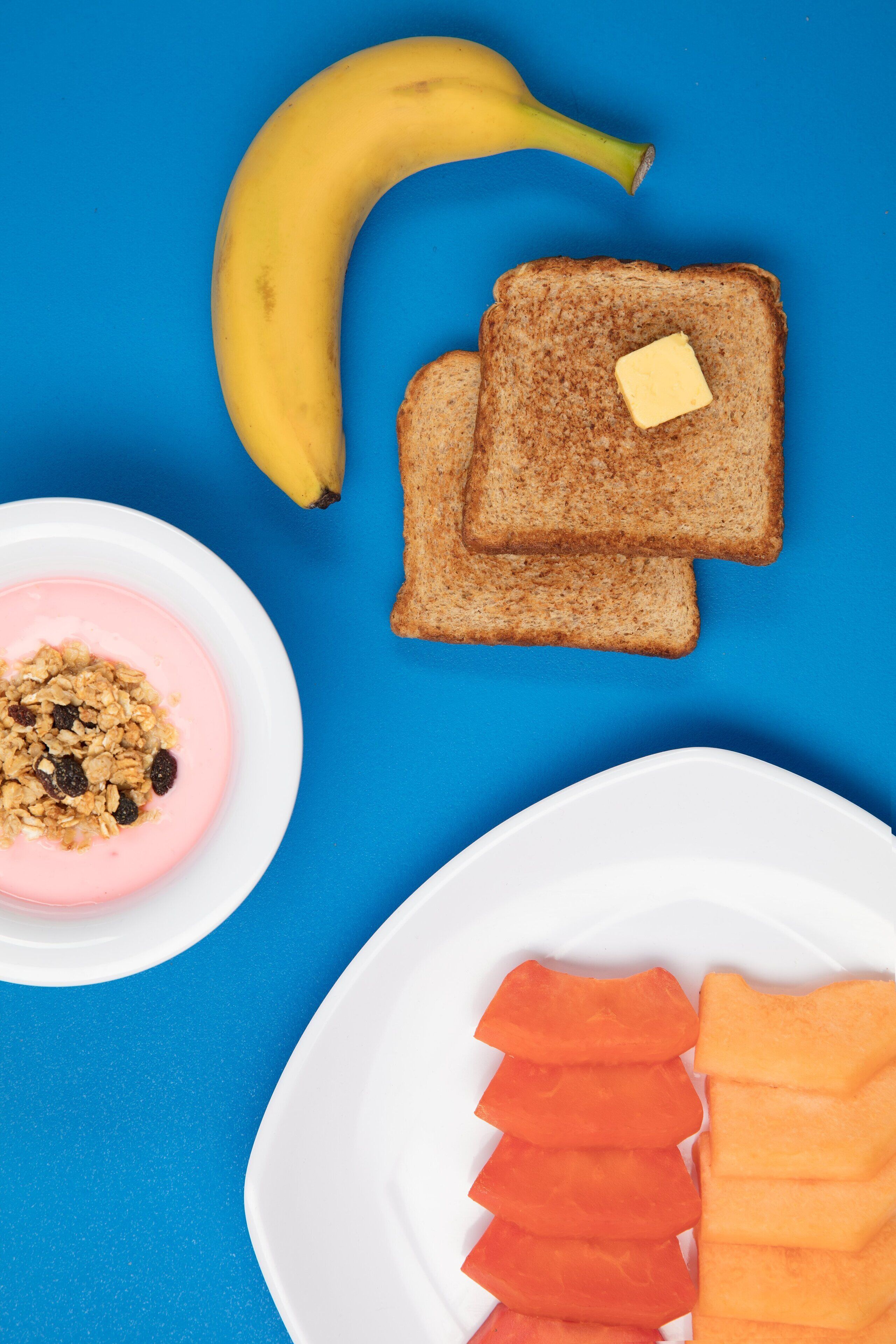 Disposição para o pequeno-almoço com torradas, banana, iogurte com granola e fruta cortada sobre uma mesa azul.