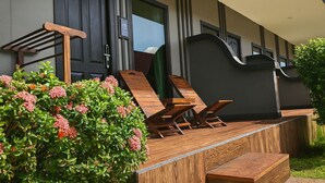 Uma área de estar moderna ao ar livre, com espreguiçadeiras de madeira e uma pequena mesa, rodeada por vegetação exuberante e uma fachada de edifício em tons escuros.