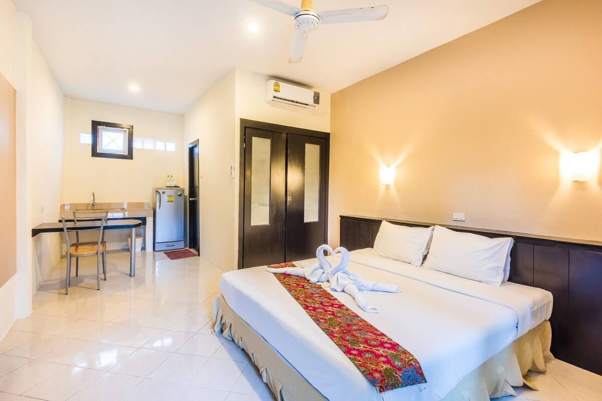Quarto de hotel com uma cama, uma escrivaninha e uma cadeira. Há um ventilador de teto, um ar condicionado e uma porta.