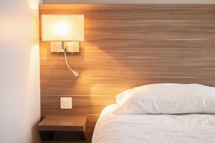 Cama bem arrumada com roupa de cama branca, cabeceira de madeira, mesa de cabeceira com abajur e tomada.