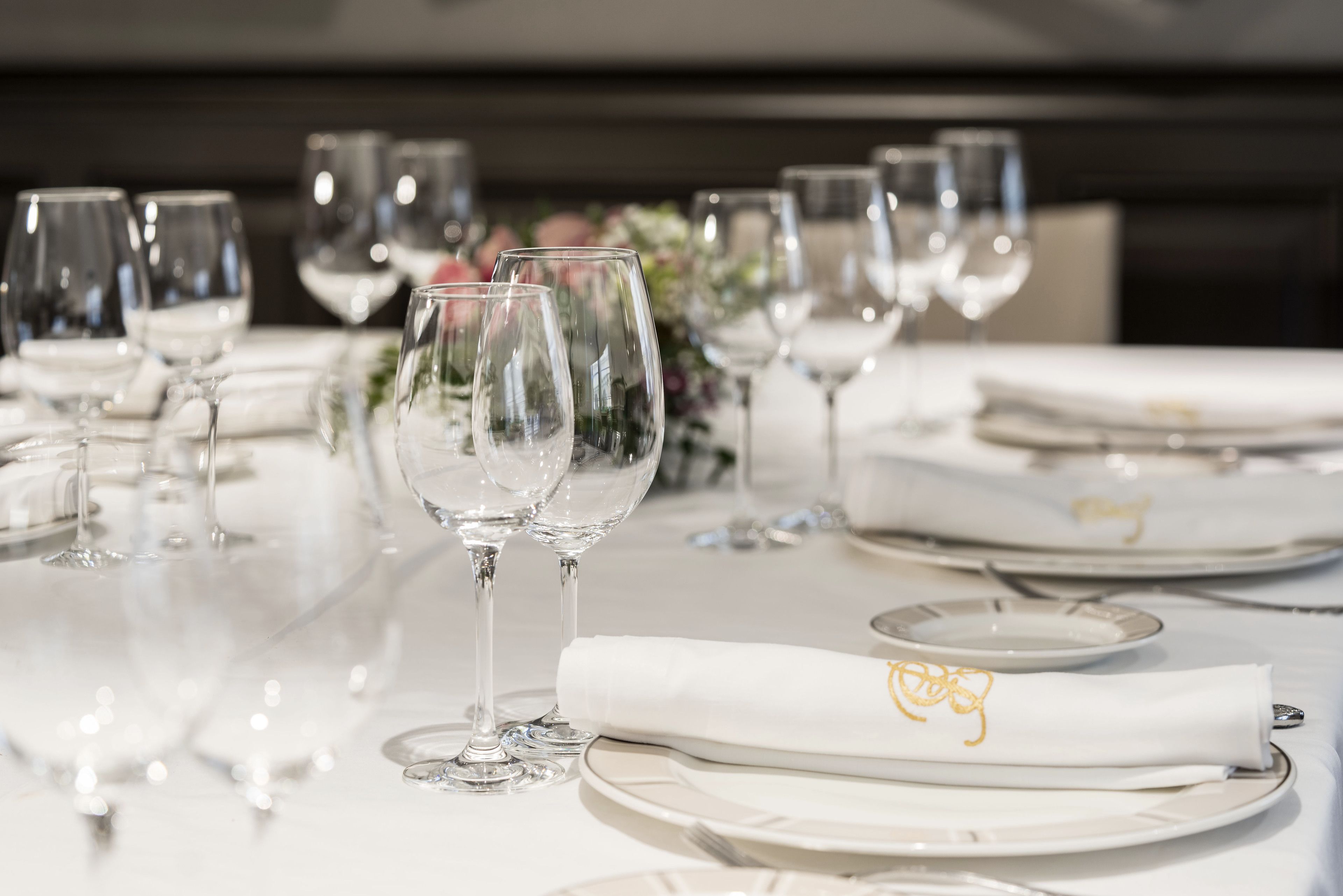 Uma mesa posta com pratos brancos, copos transparentes e guardanapos dobrados com um emblema dourado.