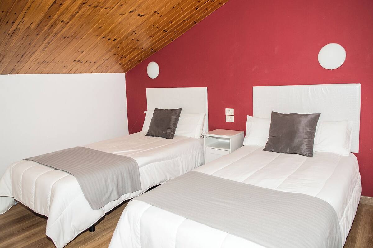 Quarto com duas camas, cada uma com cabeceira branca e travesseiro cinza. Há uma pequena mesa de cabeceira entre as camas, e o quarto tem teto de madeira.