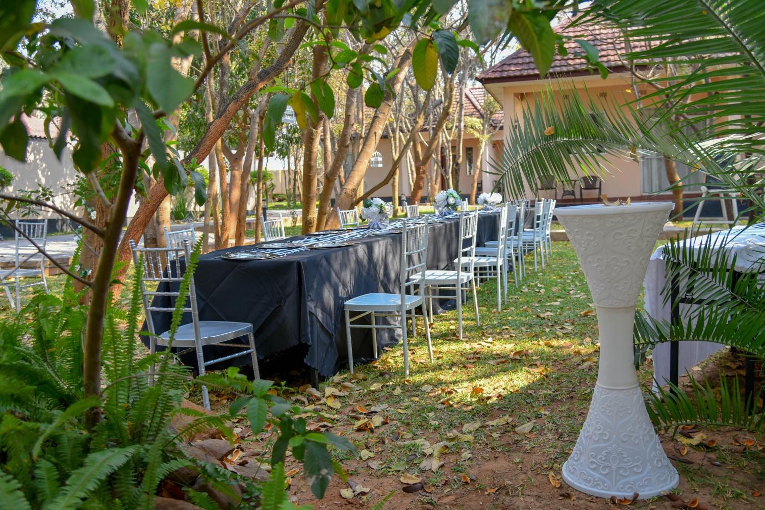 Uma área de refeições ao ar livre com mesas e cadeiras, rodeada por árvores e um vaso decorativo branco.