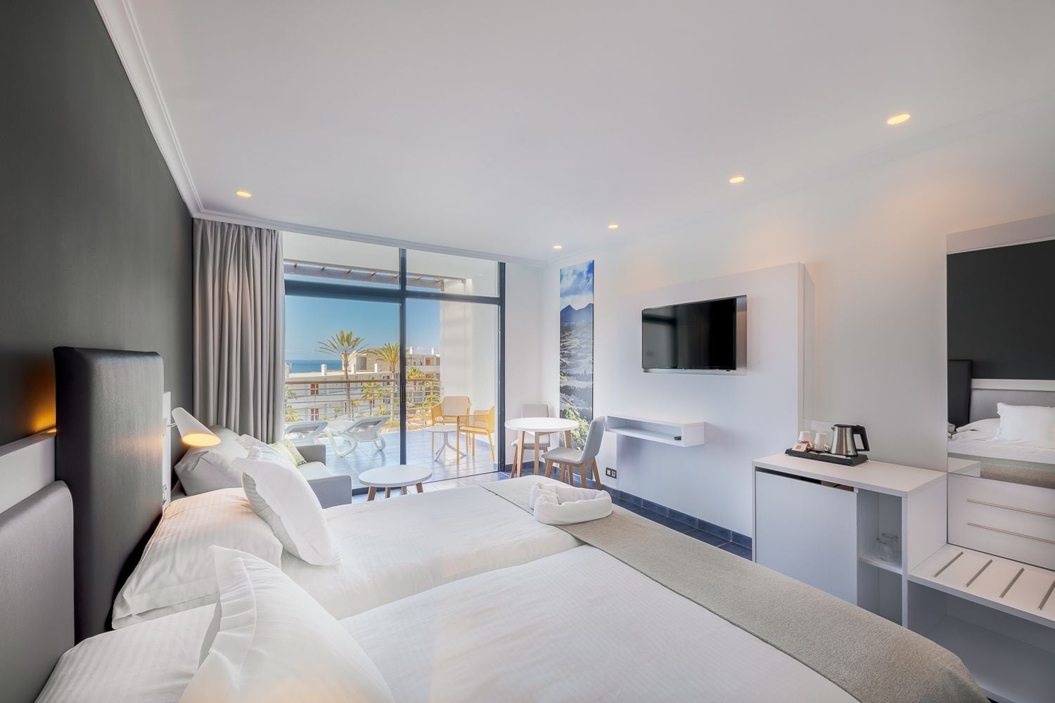 Quarto de hotel moderno com duas camas, televisão, secretária e varanda com vista para o mar.