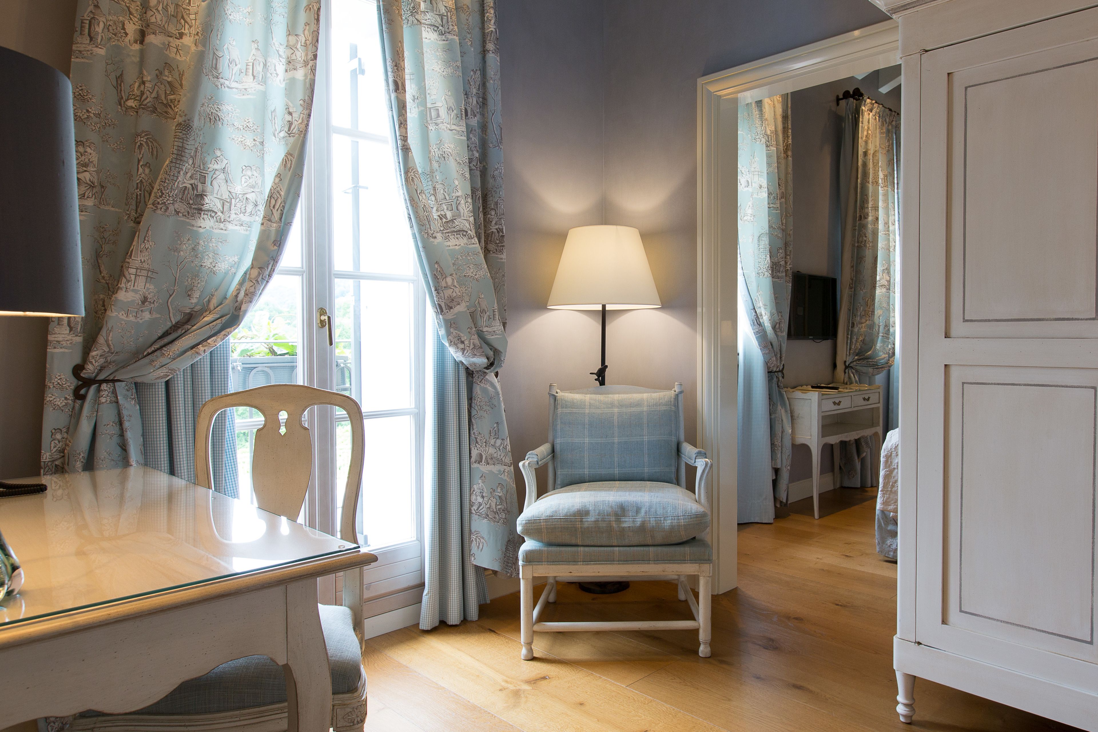 Quarto com chão de madeira, uma cadeira branca, uma mesa, um abajur e um guarda-roupa.