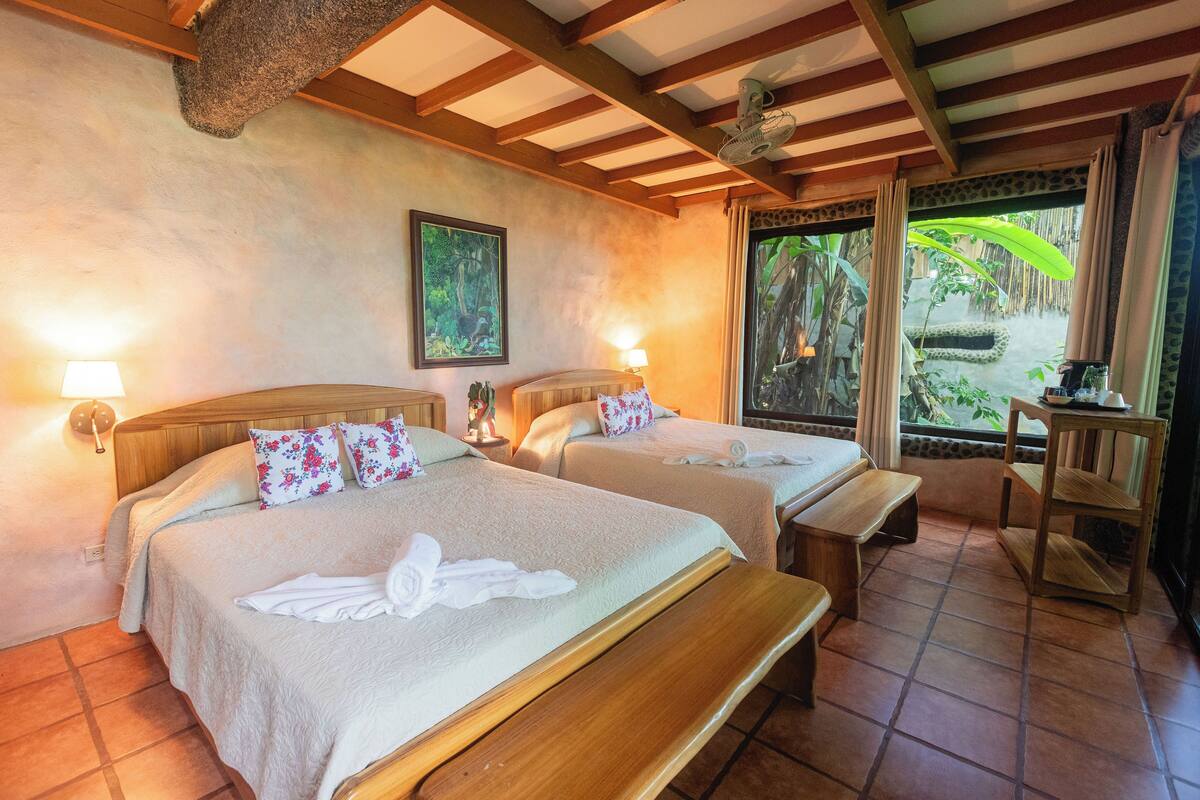 Quarto de hotel com duas camas, um banco de madeira, uma mesa pequena e vista para o jardim pela janela.
