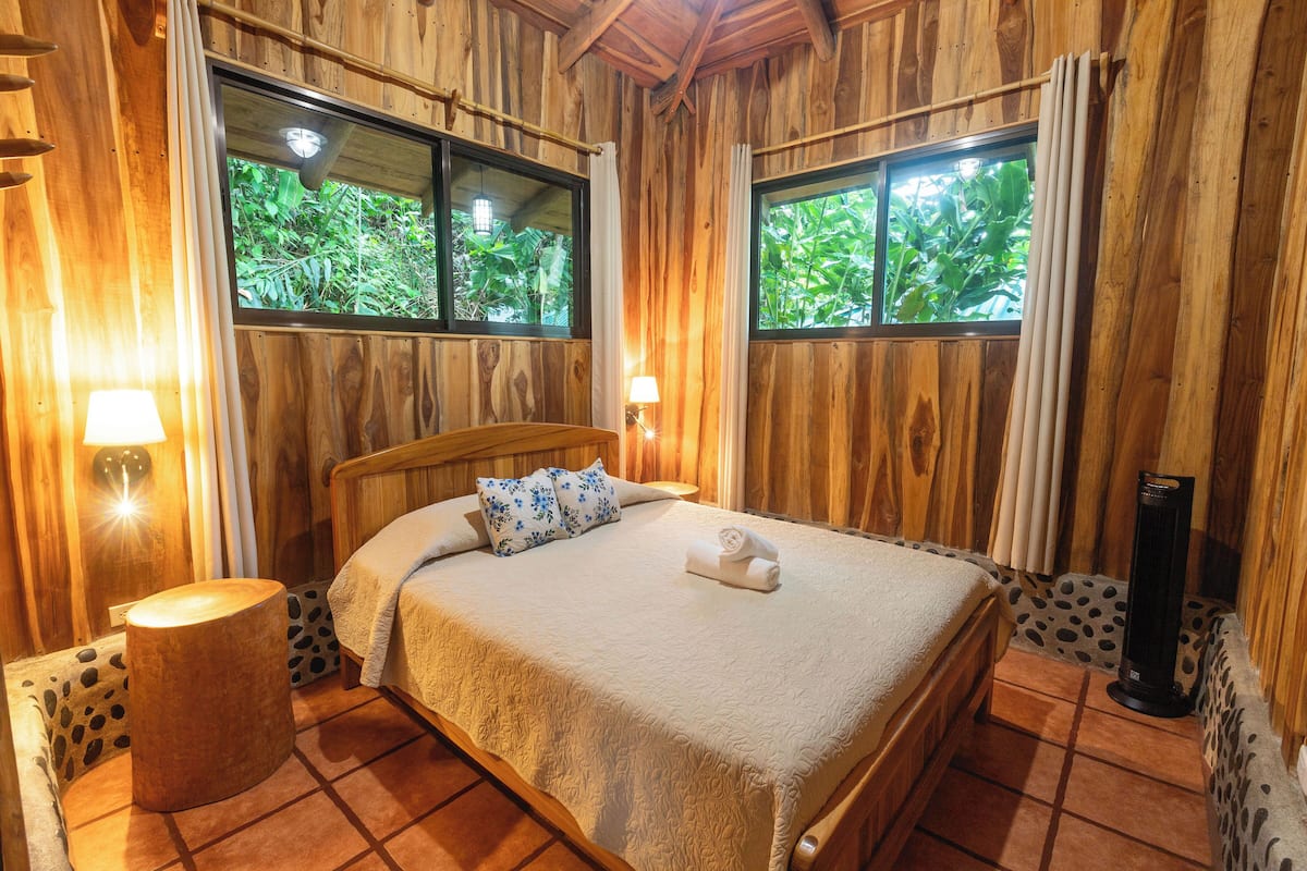 Um quarto de madeira com uma cama, duas janelas, um abajur e um aquecedor.