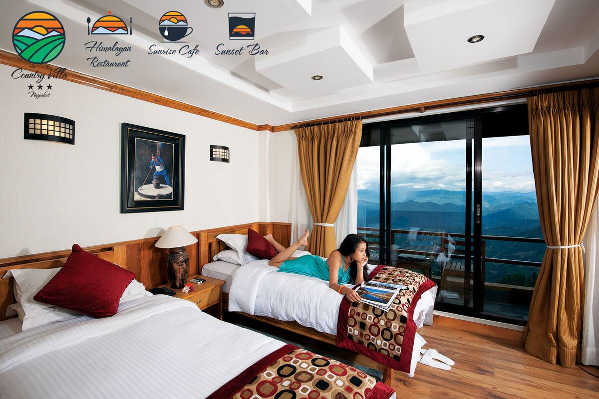 Quarto de hotel com uma grande janela que oferece vista para as montanhas, uma cama com travesseiro vermelho e uma mesa de cabeceira com abajur.