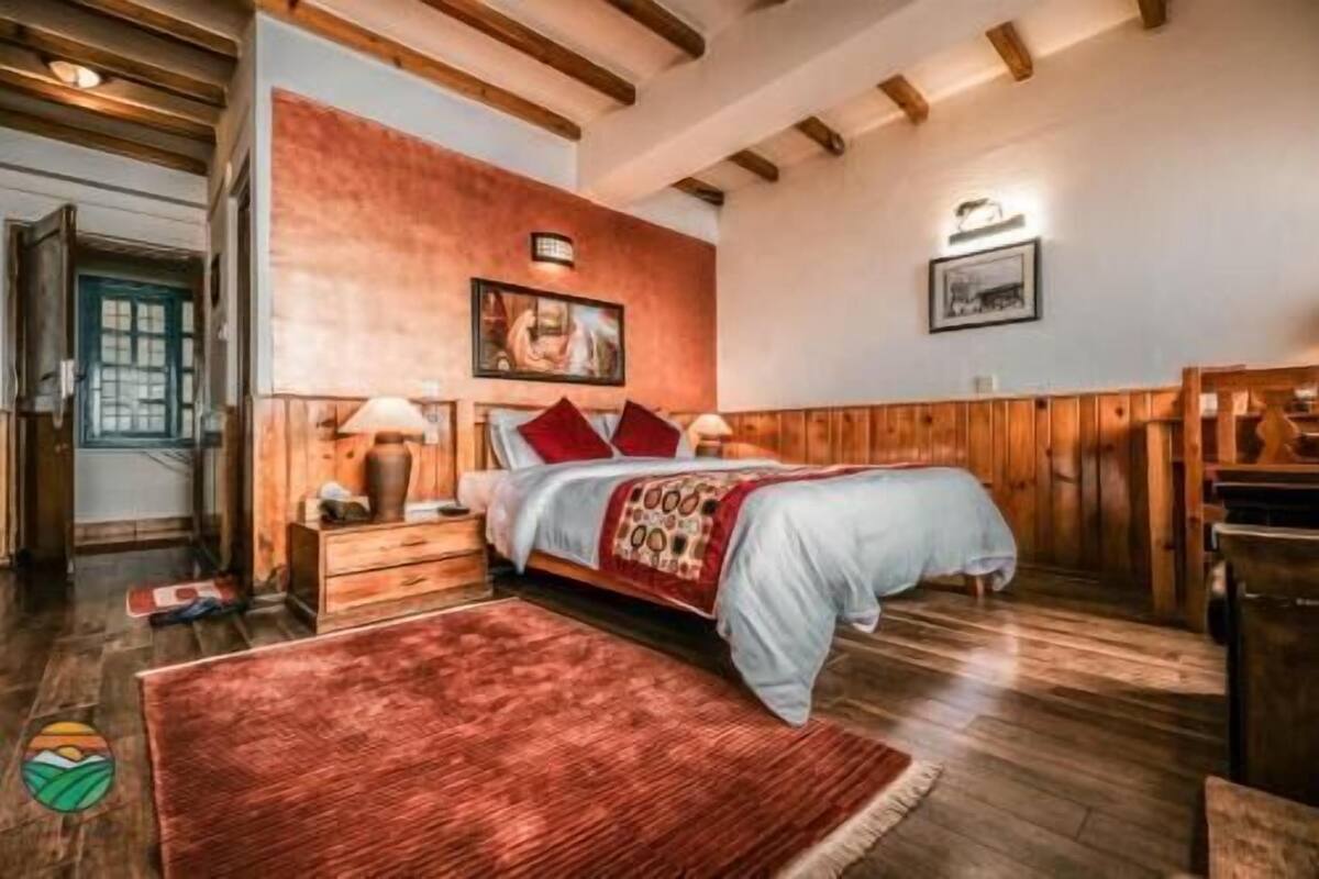 Quarto com piso de madeira, cama com colcha vermelha e branca, mesa de cabeceira com abajur e um quadro na parede.