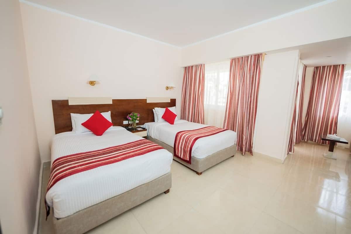 Quarto de hotel com duas camas, cada uma com almofadas vermelhas e roupa de cama listrada. Há uma mesa de cabeceira com um vaso de flores e uma luminária. O quarto tem uma janela grande com cortinas listradas e vista para o exterior.