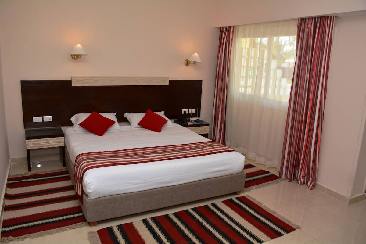 Quarto de hotel com uma cama grande, duas mesas de cabeceira, um criado-mudo, cortina listrada em vermelho e branco, e vista para o exterior.
