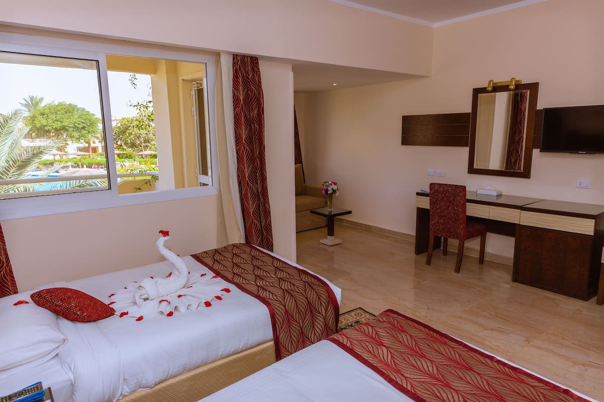 Quarto de hotel com duas camas, uma escrivaninha, uma cadeira, televisão e janela com vista para a piscina e áreas verdes.