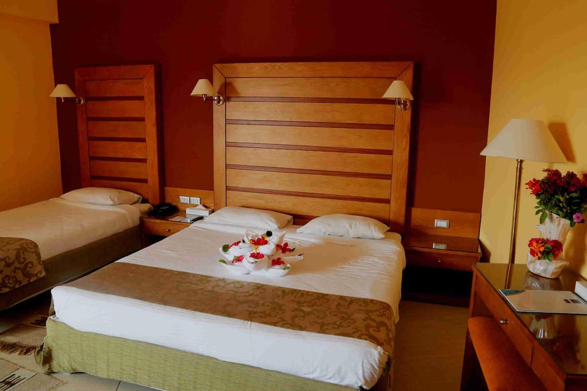 Quarto de hotel com duas camas de solteiro, cabeceira de madeira, mesas de cabeceira com abajures, escrivaninha com cadeira e um vaso com flores.