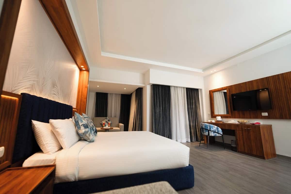 Quarto de hotel moderno com uma cama grande, uma escrivaninha, uma televisão e um banheiro visível através de uma divisória de vidro.