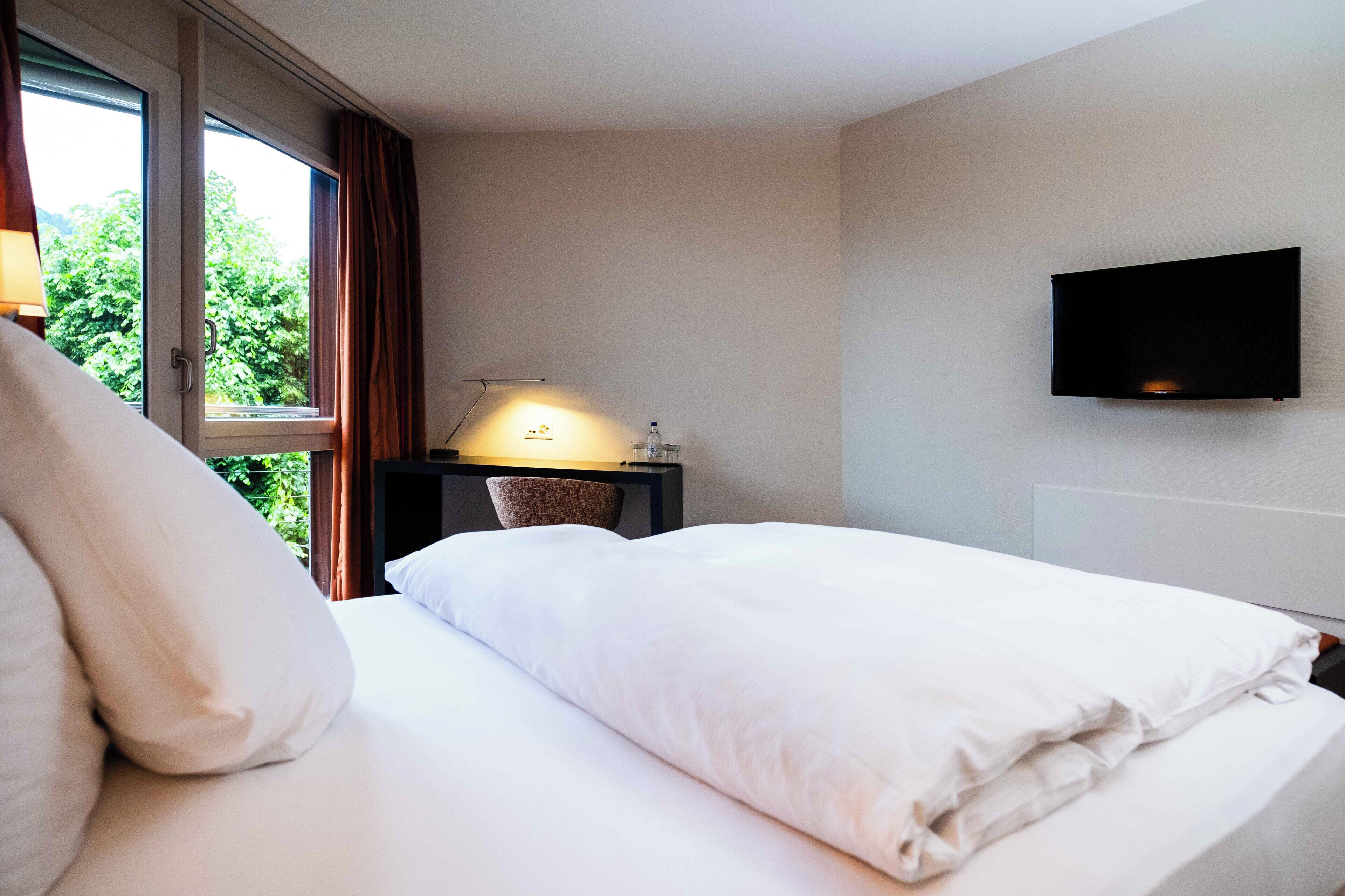 Quarto de hotel com cama, escrivaninha com luminária, TV fixada na parede e janela com cortinas.