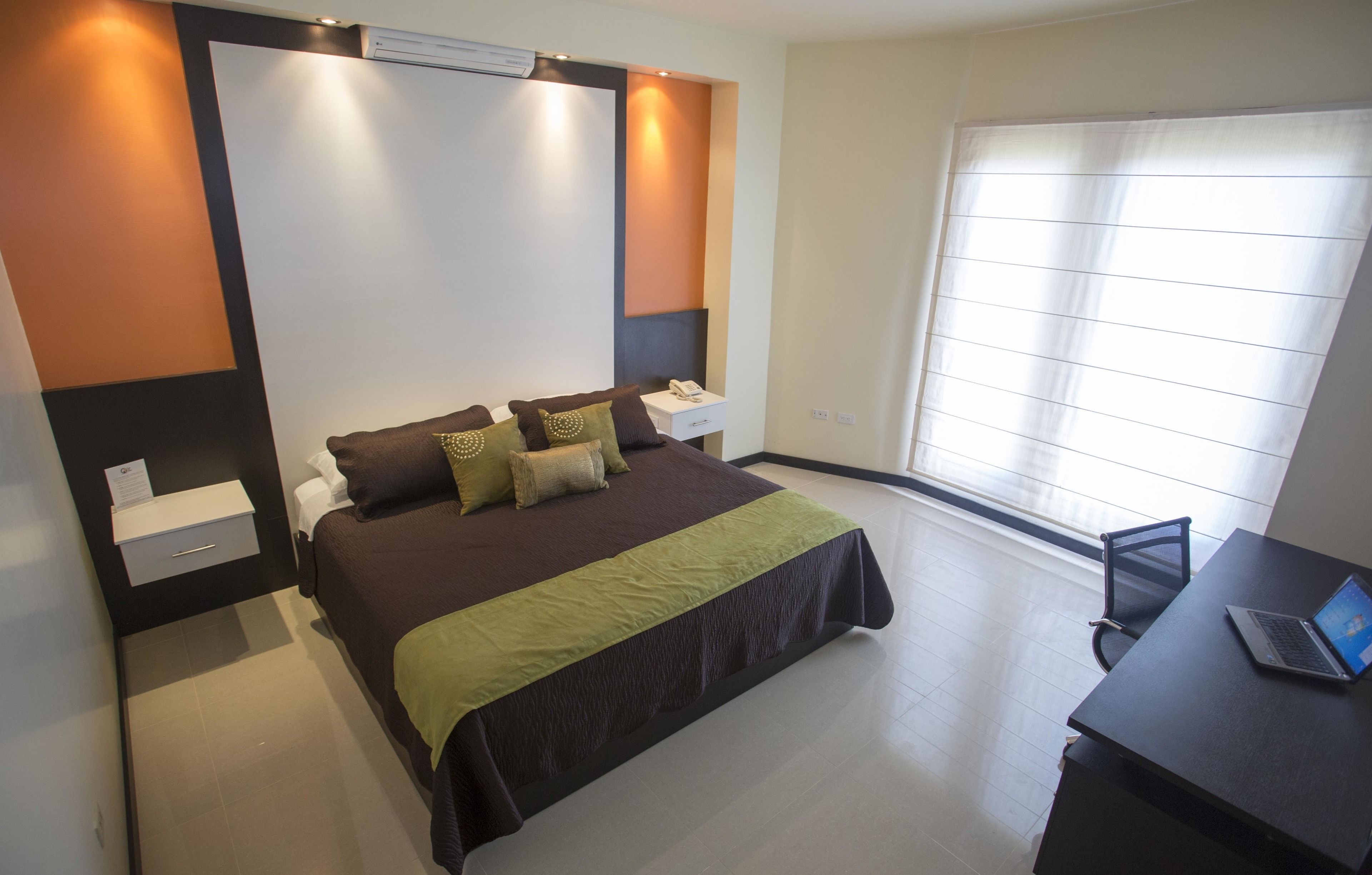 Quarto de hotel moderno com uma cama grande, mesa de cabeceira e escrivaninha com um laptop.