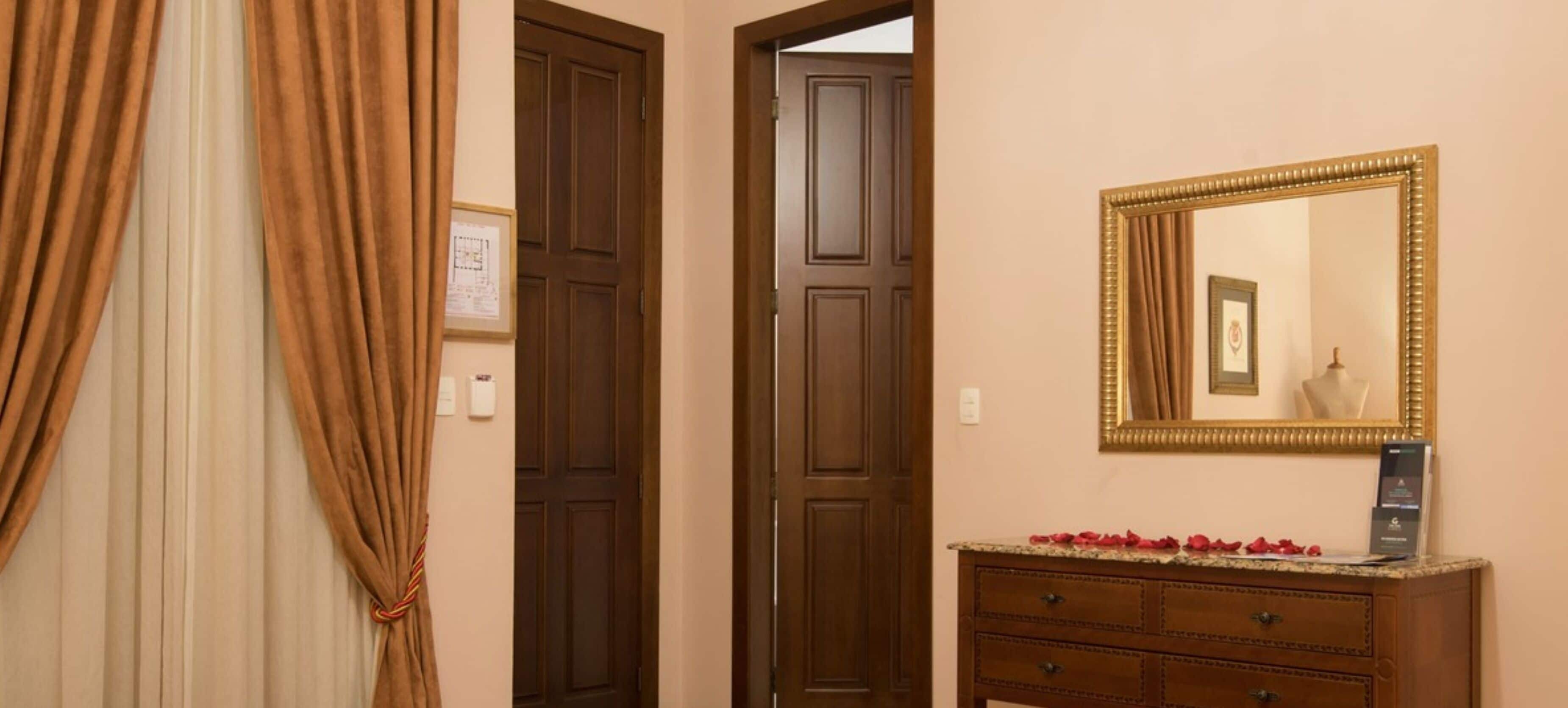 Quarto de hotel com porta de madeira, espelho com moldura dourada e cômoda com flores vermelhas.