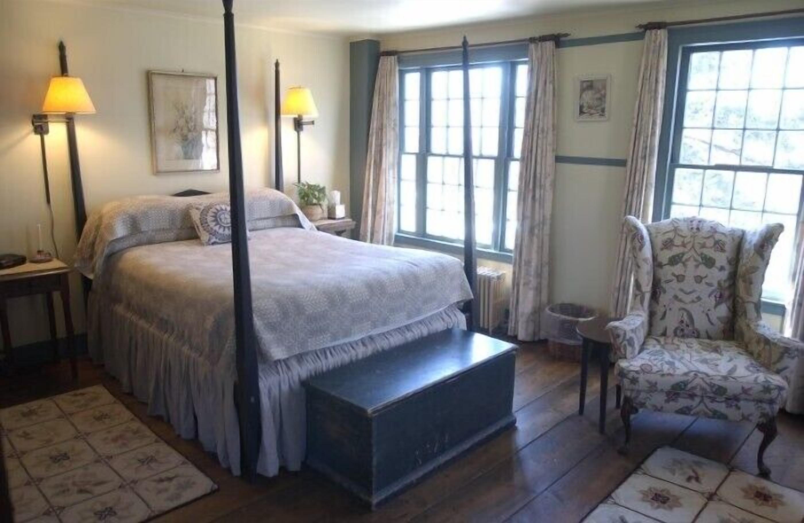 Uma cama com dossel, um banco, uma poltrona florida e vista para o exterior por grandes janelas.