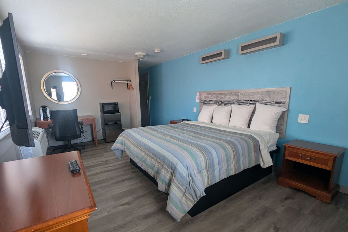 Quarto com cama, escrivaninha, cadeira, micro-ondas, geladeira e uma pequena mesa.