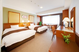 Quarto de hotel com duas camas, uma escrivaninha, uma cadeira, uma mesinha, uma planta e uma janela grande com cortinas.