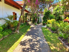 Uma trilha pavimentada ladeada por vegetação exuberante e plantas floridas, conduzindo a um edifício com telhado de azulejos e parede branca.