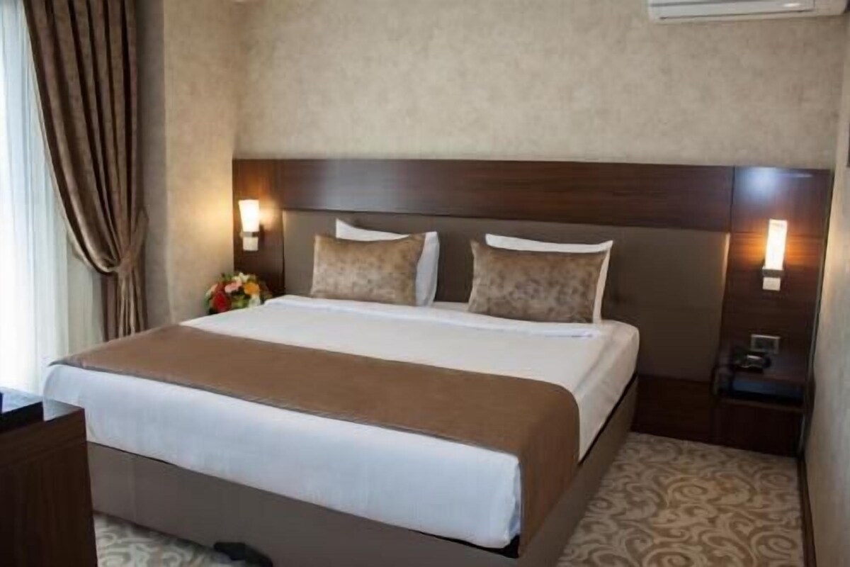 Quarto de hotel com cama grande, cabeceira, mesas de cabeceira e apoio para objetos.